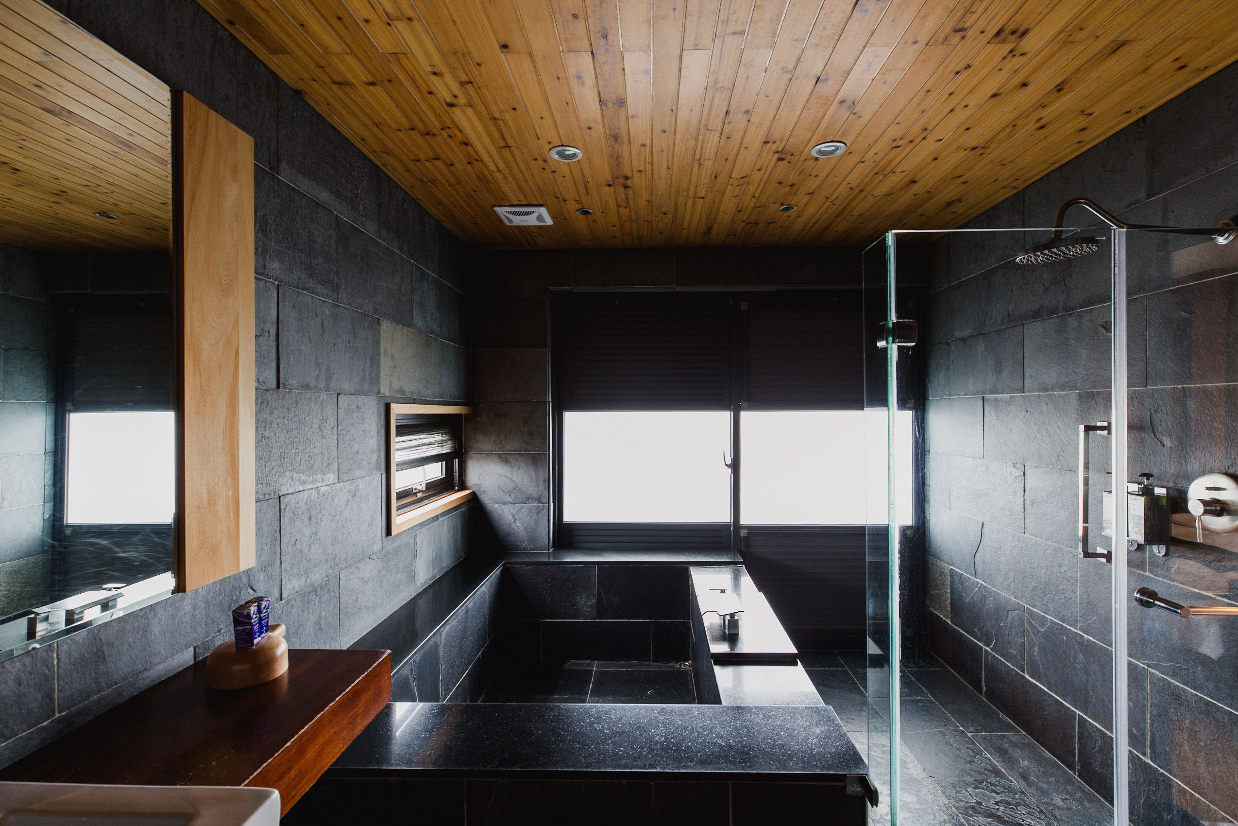 Casa de banho moderna com teto de madeira, cabine de chuveiro em vidro e uma janela grande que permite entrada de luz natural.