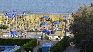 Uma praia com fileiras de cadeiras amarelas e guarda-sóis azuis, céu claro e um veleiro ao longe.