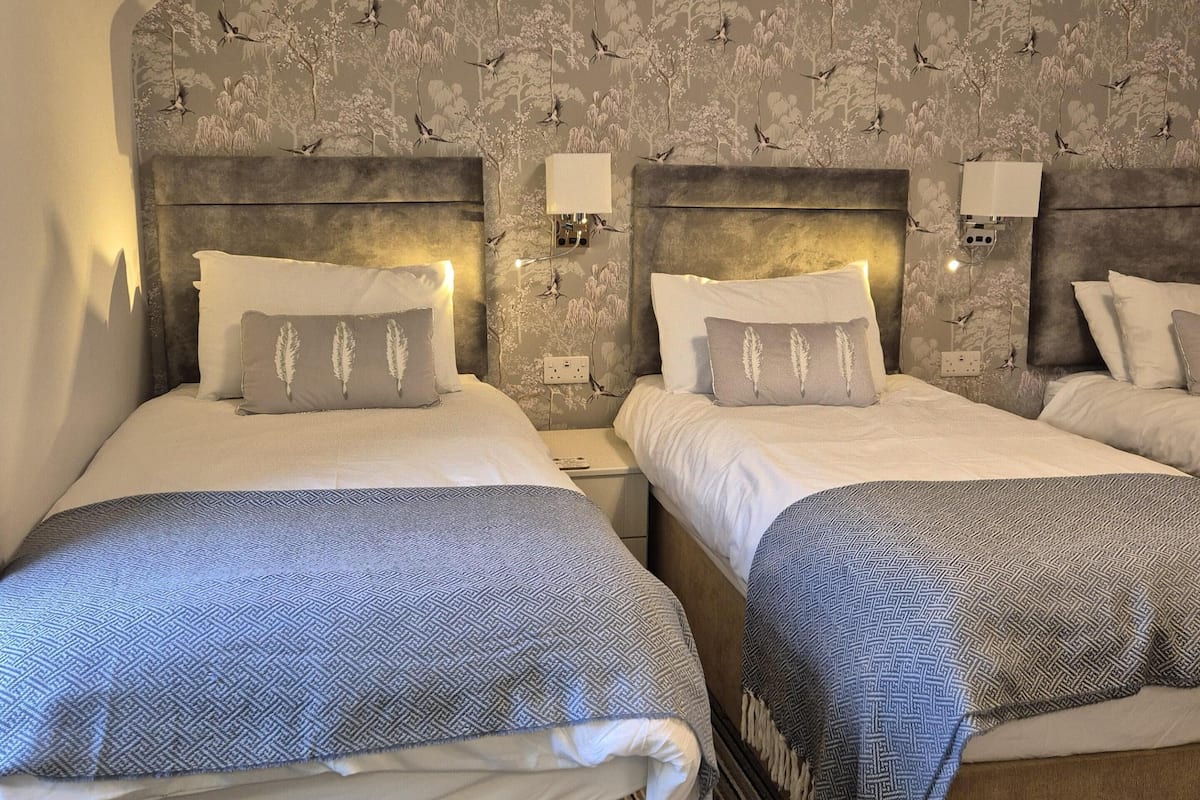 Quarto de hotel com duas camas, cada uma com colcha estampada, almofadas brancas e cabeceira com papel de parede floral. Há luminárias de cabeceira e uma tomada na parede.