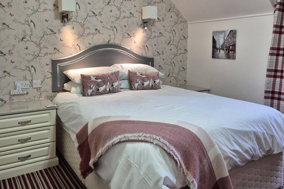 Quarto com cama, mesas de cabeceira, luminária na parede e uma pintura.