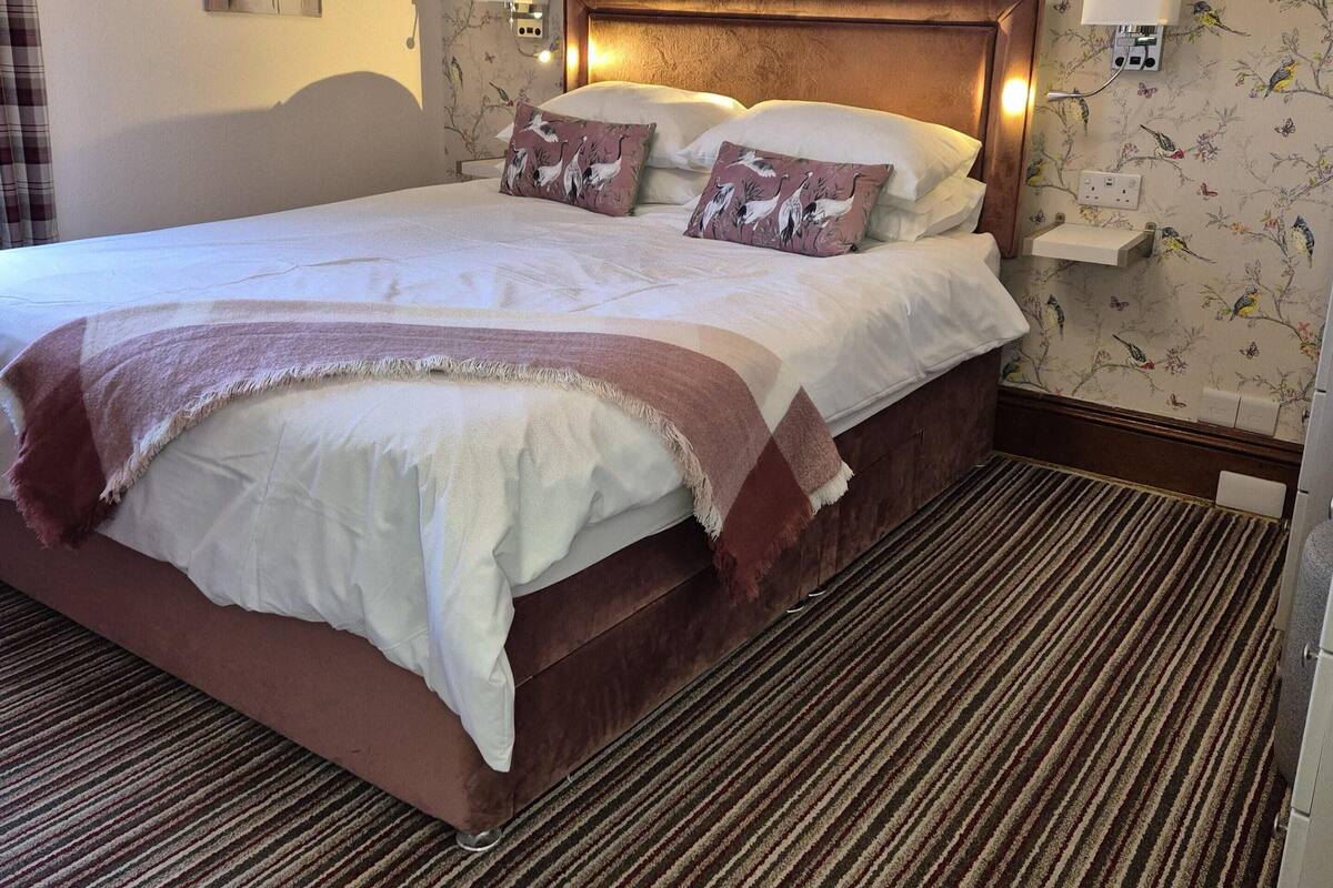 Quarto de hotel com uma cama grande, duas luminárias fixadas na parede, papel de parede floral e um quadro na parede.