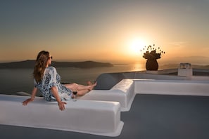 Uma mulher relaxa em um banco acolchoado de branco, apreciando a vista panorâmica do mar e do pôr do sol.