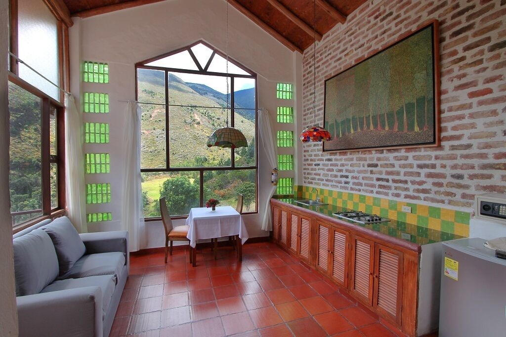 Quarto com uma grande janela, uma mesa com cadeiras, um sofá, cozinha com fogão e um quadro na parede.