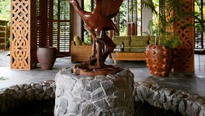 Escultura de madeira de um pássaro sobre um pedestal de pedra, cercada por uma cerca de madeira com entalhes elaborados, em um quarto com um sofá verde e uma planta em vaso.