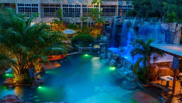 Piscina de resort com cachoeira, cercada por plantas tropicais e palmeiras.