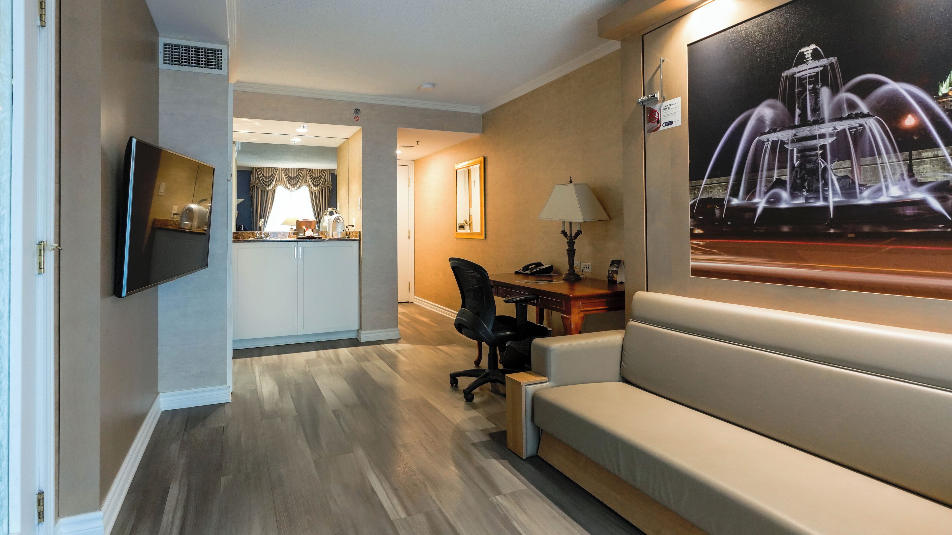 Quarto de hotel com televisão de tela plana na parede, cozinha compacta com pia e bancada, escrivaninha com cadeira e um pôster grande de uma fonte.