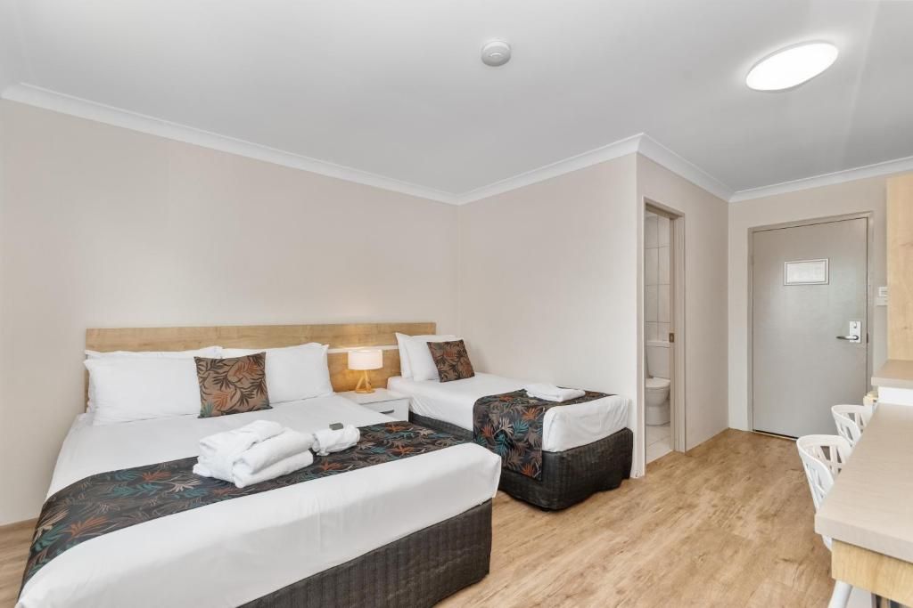 Quarto de hotel com duas camas, cabeceira de madeira, uma pequena escrivaninha e porta de banheiro.