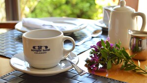 Uma mesa posta com uma xícara branca identificada com "acropol hotel HALANDRI", um bule branco, uma colher de prata e um pequeno vaso com flores roxas.