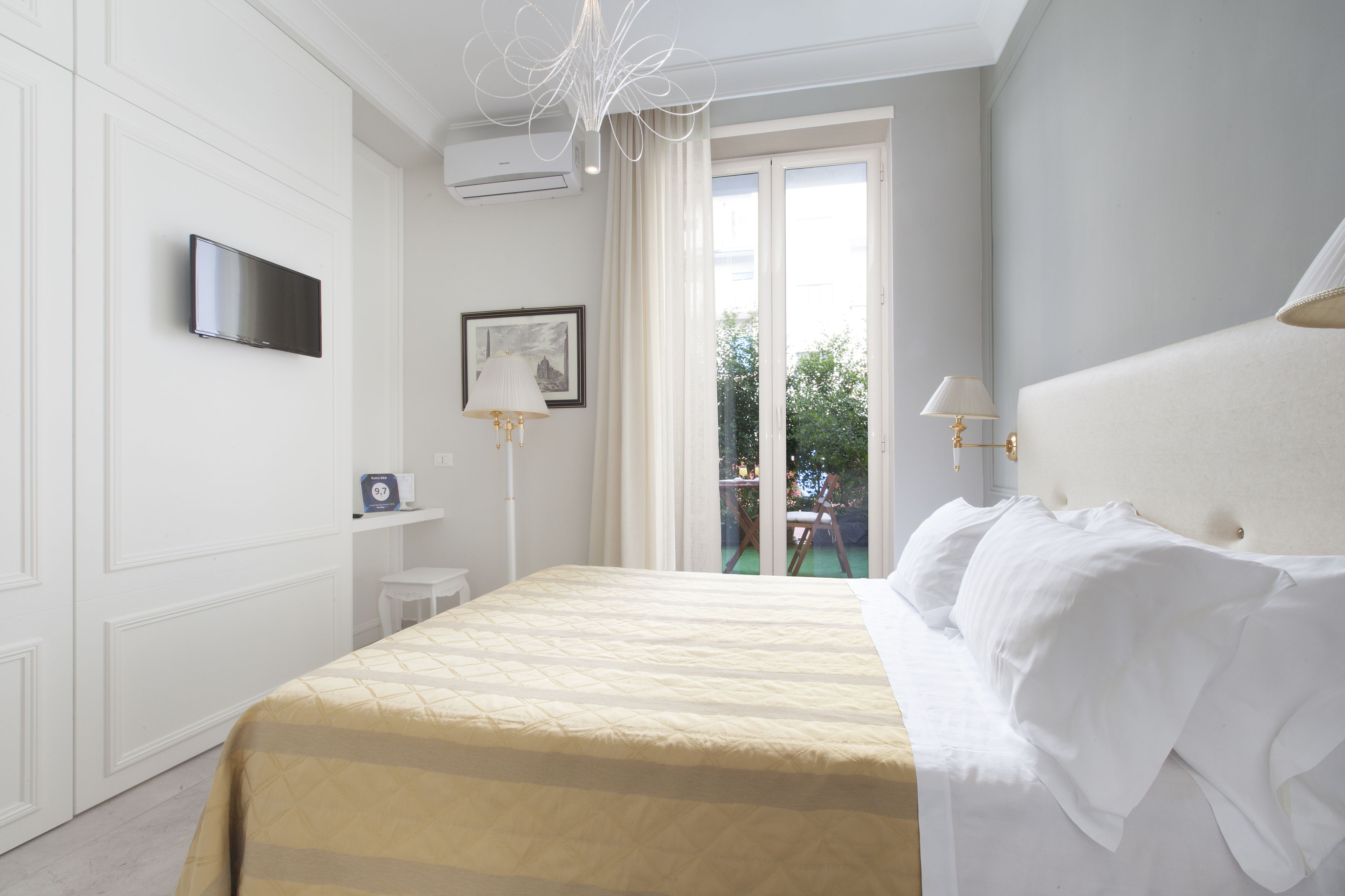 Quarto com cama, televisão na parede, janela com cortinas e varanda com mesa e cadeiras.
