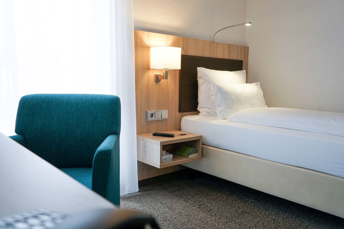 Quarto de hotel com cama, escrivaninha, cadeira, mesa de cabeceira com abajur e uma pequena prateleira com um controle remoto e um objeto verde.