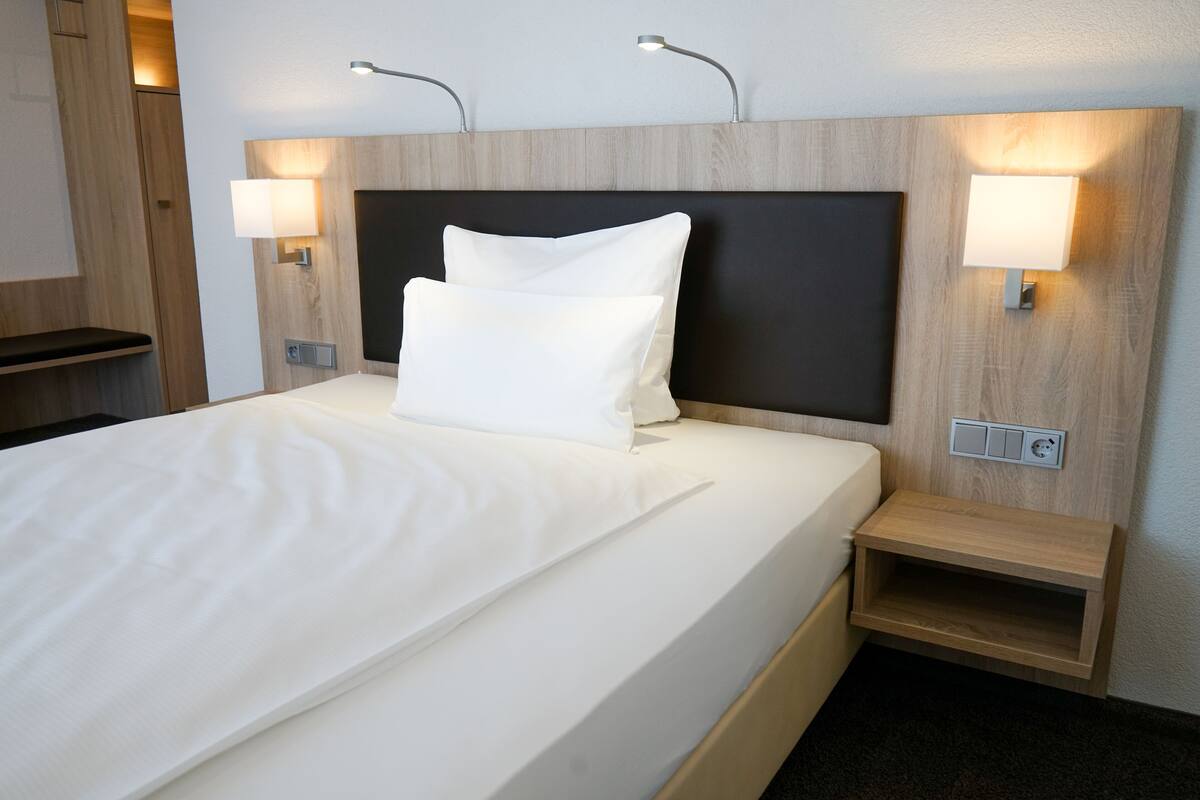 Cama bem arrumada com lençóis brancos e duas almofadas, cabeceira de madeira e abajures laterais.