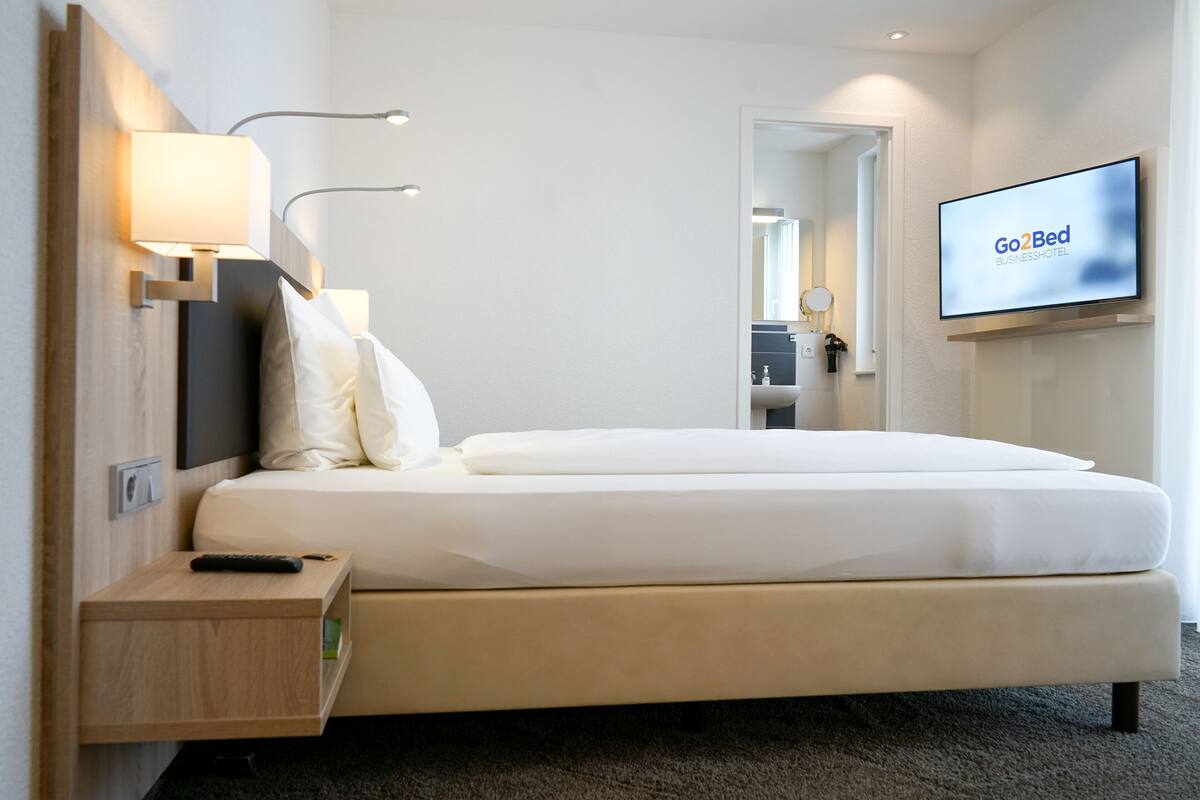 Quarto de hotel moderno com uma cama grande, luminárias de cabeceira, uma TV e uma mesa de cabeceira de madeira.