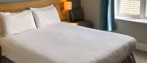 Cama bem arrumada com roupa de cama branca, cabeceira de madeira, luminária de cabeceira e janela com cortinas.