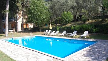 Uma piscina retangular com borda de pedra, rodeada por espreguiçadeiras brancas e um guarda-sol amarelo.