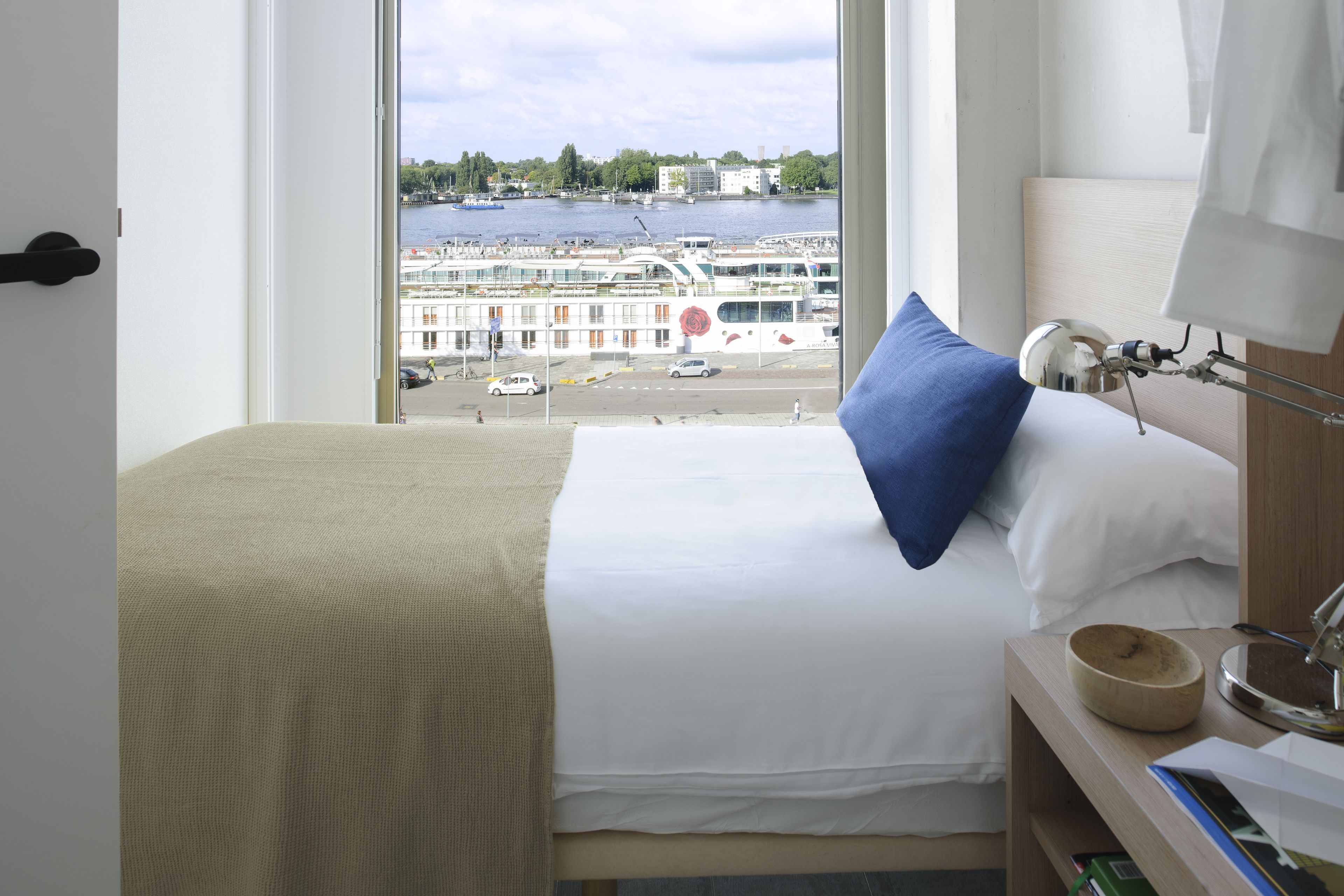 Quarto de hotel com uma cama, vista para um rio e edifícios, mesa de cabeceira com abajur e janela com vista para a cidade.