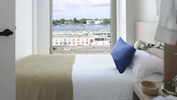 Quarto de hotel com uma cama, vista para um rio e edifícios, mesa de cabeceira com abajur e janela com vista para a cidade.