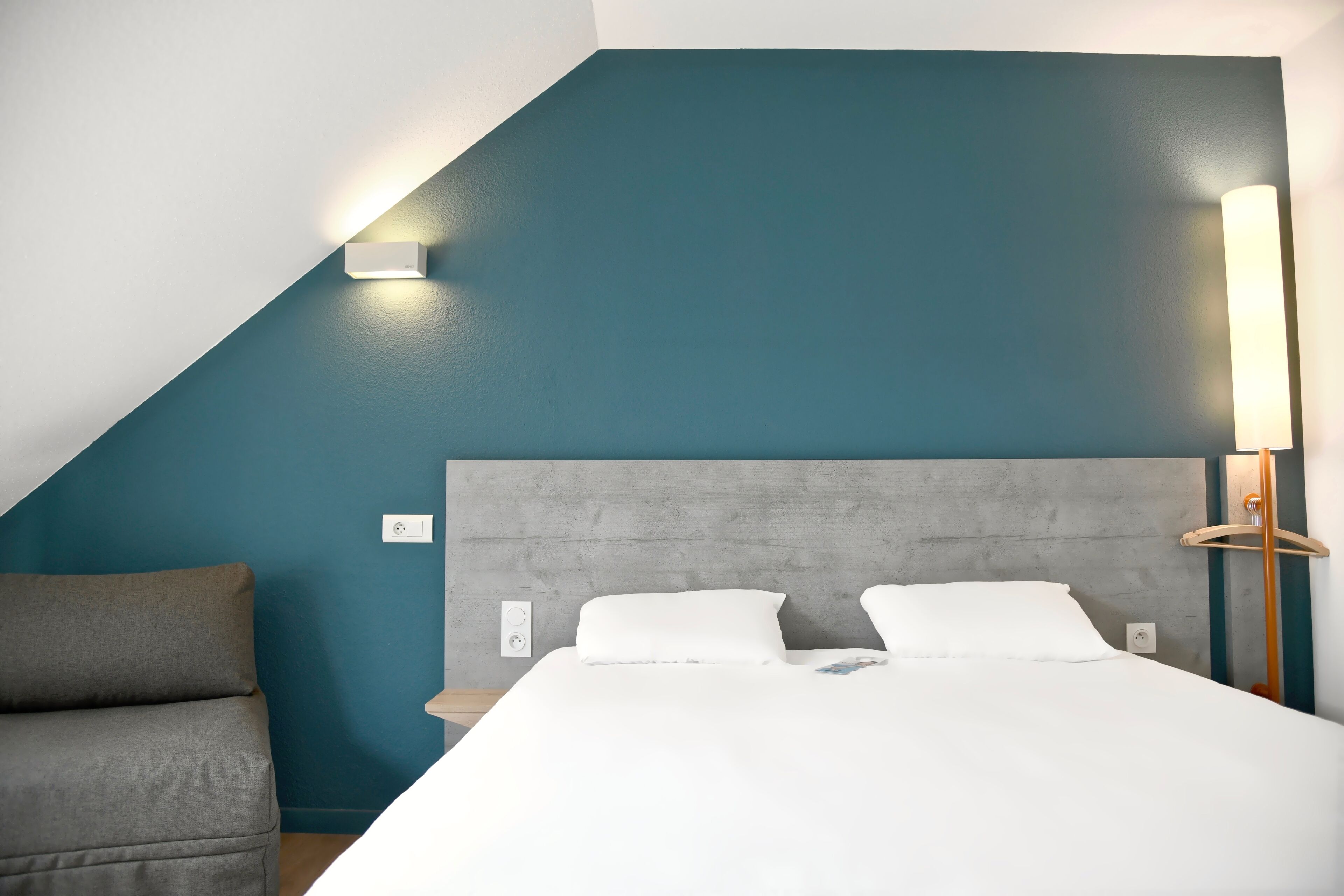 Quarto de hotel com uma cama, uma cadeira e uma luminária fixada na parede.