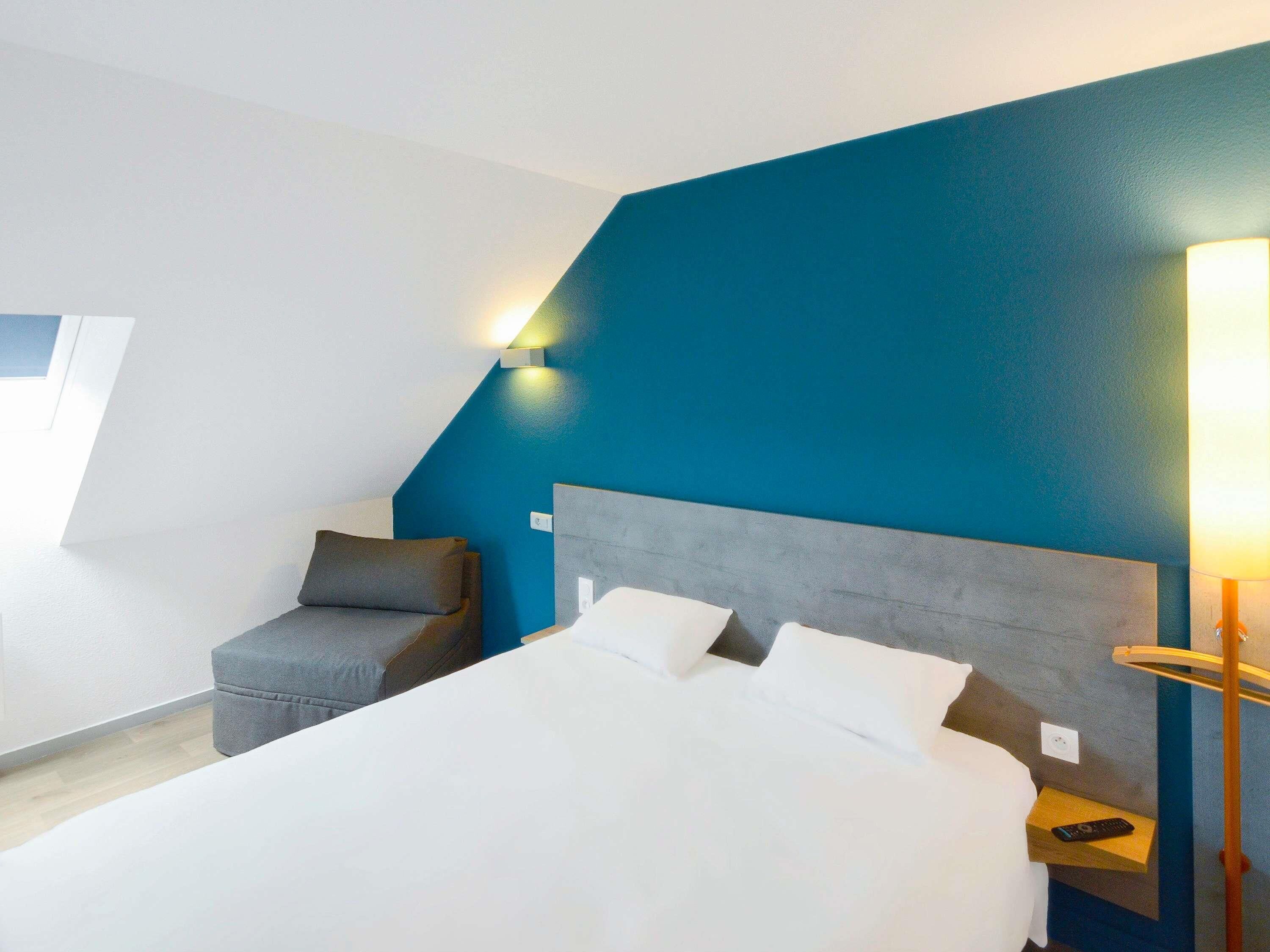 Quarto de hotel com uma cama, um pequeno sofá, uma mesa de cabeceira e uma luminária.