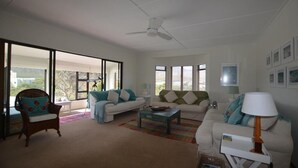 Uma sala de estar espaçosa, com grandes janelas, um ventilador de teto e diversas opções de assentos, incluindo uma cadeira de vime, dois sofás brancos e um tapete colorido.