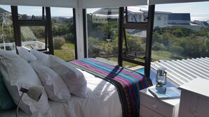 Quarto com cama grande, roupa de cama branca, manta listrada colorida e mesa de cabeceira com um candeeiro.