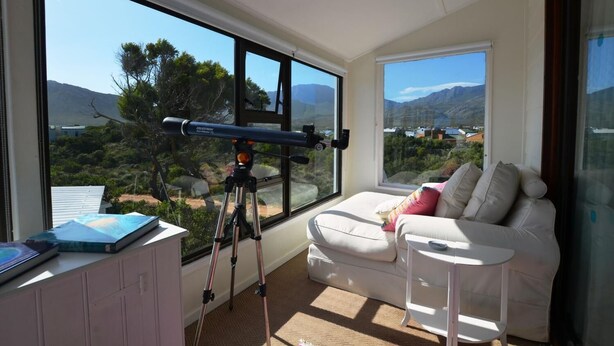 Um quarto com um telescópio sobre um tripé, um sofá branco com almofadas e janelas amplas com vista para uma paisagem montanhosa.