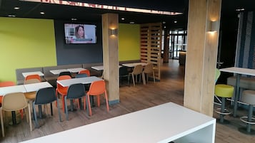 Interior moderno de restaurante com mesas brancas, cadeiras coloridas e uma grande tela exibindo uma programação.