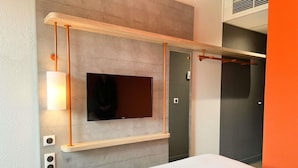 Quarto de hotel moderno com televisão montada, mesa de cabeceira e guarda-roupa embutido.