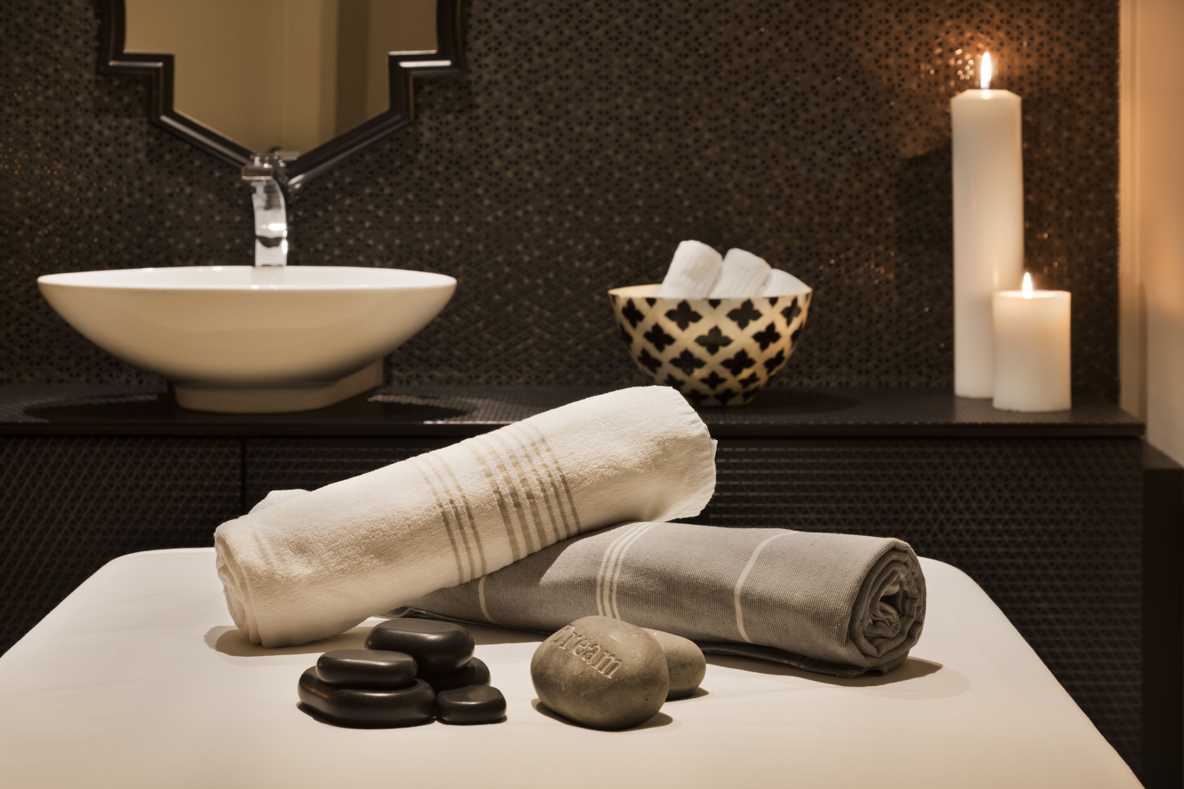 Uma área de spa com uma pia branca, uma tigela com toalhas dobradas, uma vela acesa e uma mesa para massagem de pedra.