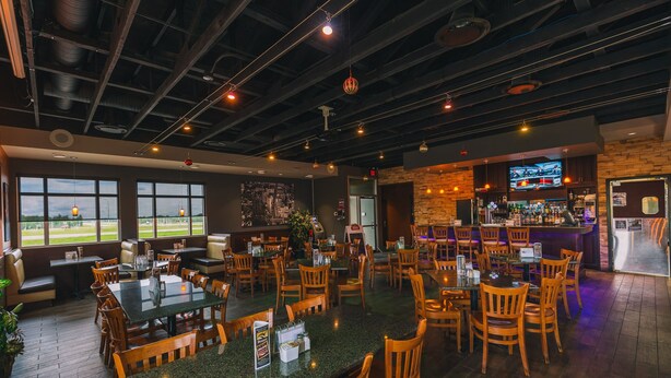 Restaurante com várias áreas de estar, incluindo cabines e mesas. Há um bar com uma tela de TV e um balcão. O restaurante possui janelas amplas e vista para o exterior.