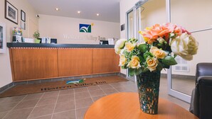 Uma área de recepção com balcão de madeira, um logotipo na parede e um arranjo floral sobre uma mesa.
