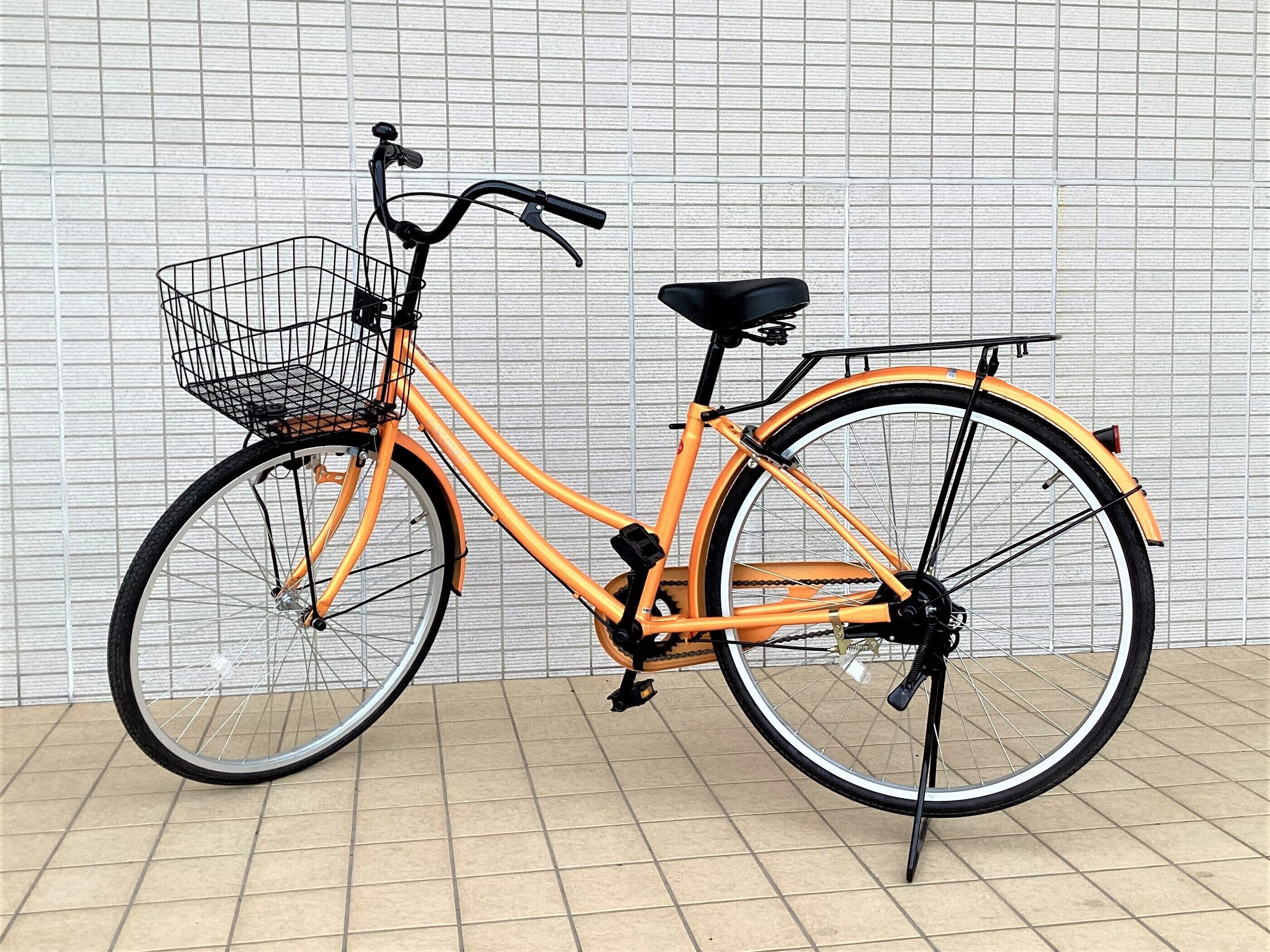 Uma bicicleta laranja estacionada contra uma parede de azulejos, com um cesto frontal e um assento.