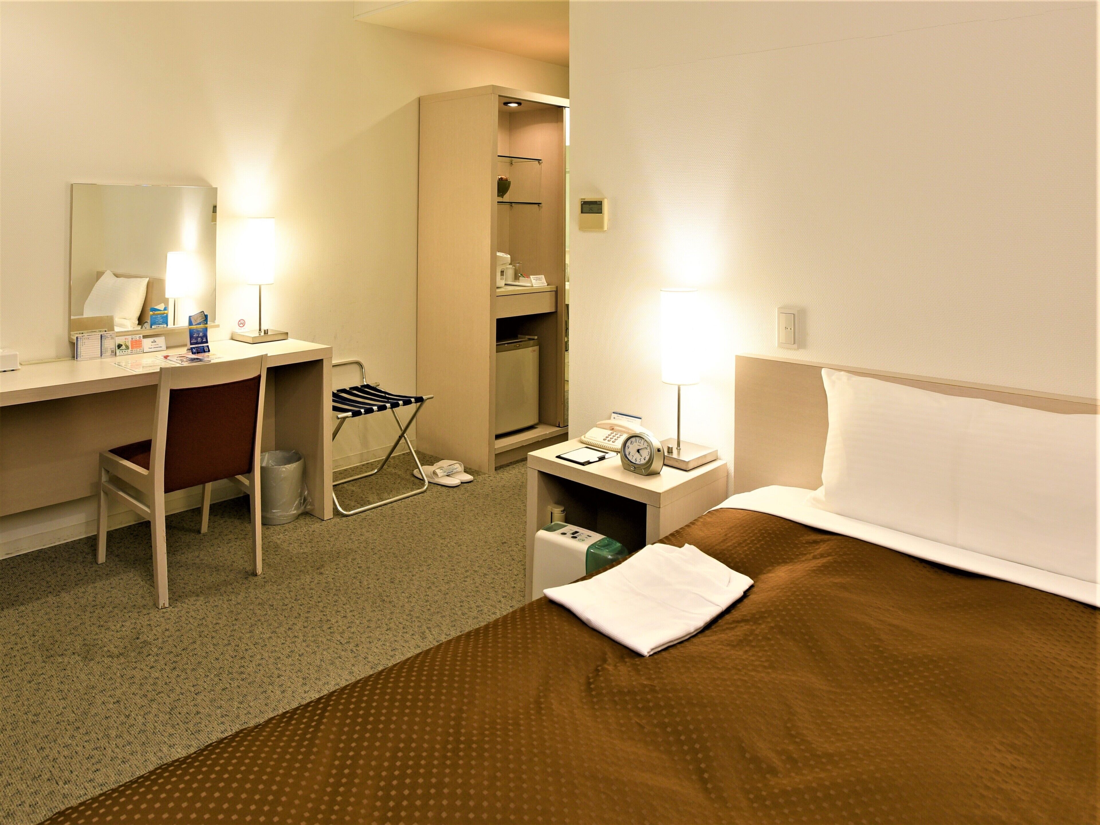 Quarto de hotel com cama, escrivaninha, cadeira, espelho, luminária, relógio e lixeira.