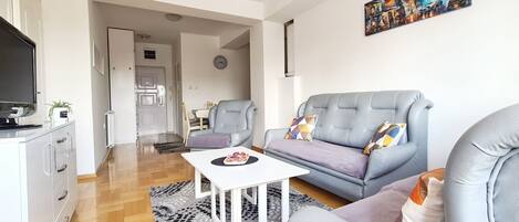 Uma sala de estar moderna com um sofá cinzento, uma mesa de centro branca e uma área de jantar ao fundo.