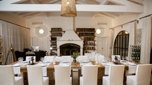 Uma sala de jantar com uma longa mesa de madeira posta para refeição, cercada por cadeiras estofadas brancas.