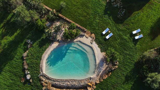 Vista aérea de uma praia com uma piscina azul límpida rodeada por vegetação e uma parede de pedra. Há espreguiçadeiras e um pequeno edifício nas proximidades.