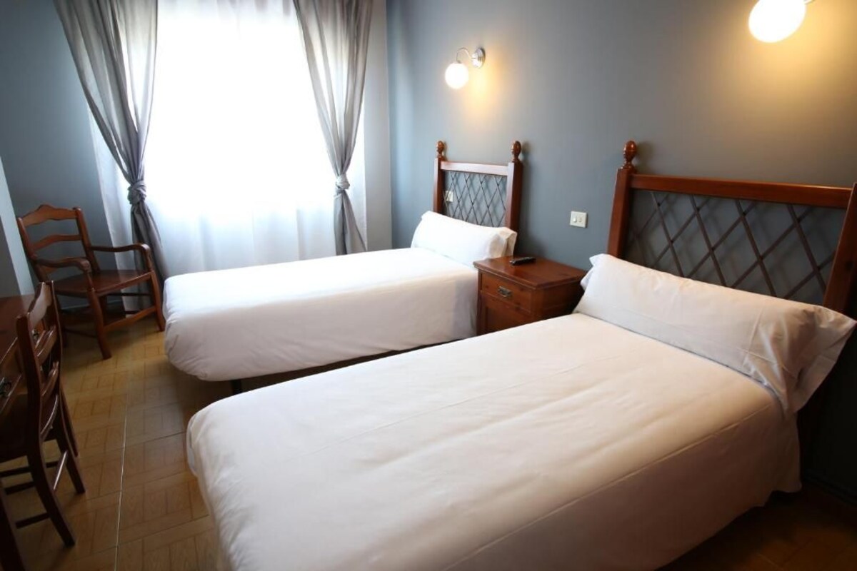 Quarto de hotel com duas camas de solteiro, uma mesinha de cabeceira de madeira, uma cadeira e uma escrivaninha.
