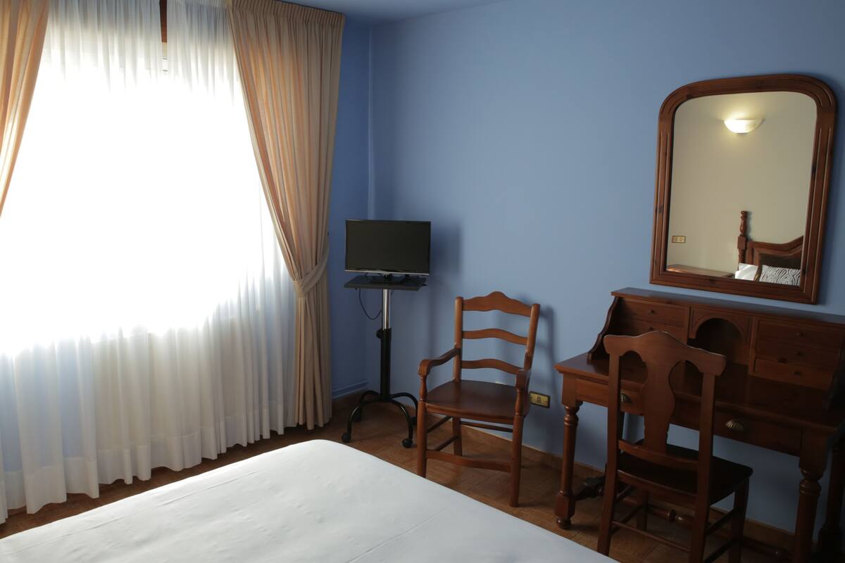 Quarto de hotel com cama, escrivaninha com cadeira, televisão em suporte, espelho e janela com cortinas.
