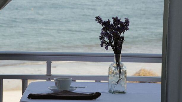 Uma mesa com toalha branca, um vaso de vidro transparente com flores roxas, uma xícara e pires brancos e um guardanapo preto.