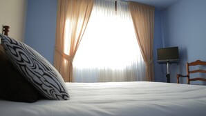 Quarto com cama, televisão sobre suporte, cadeira de madeira e janela com cortinas.