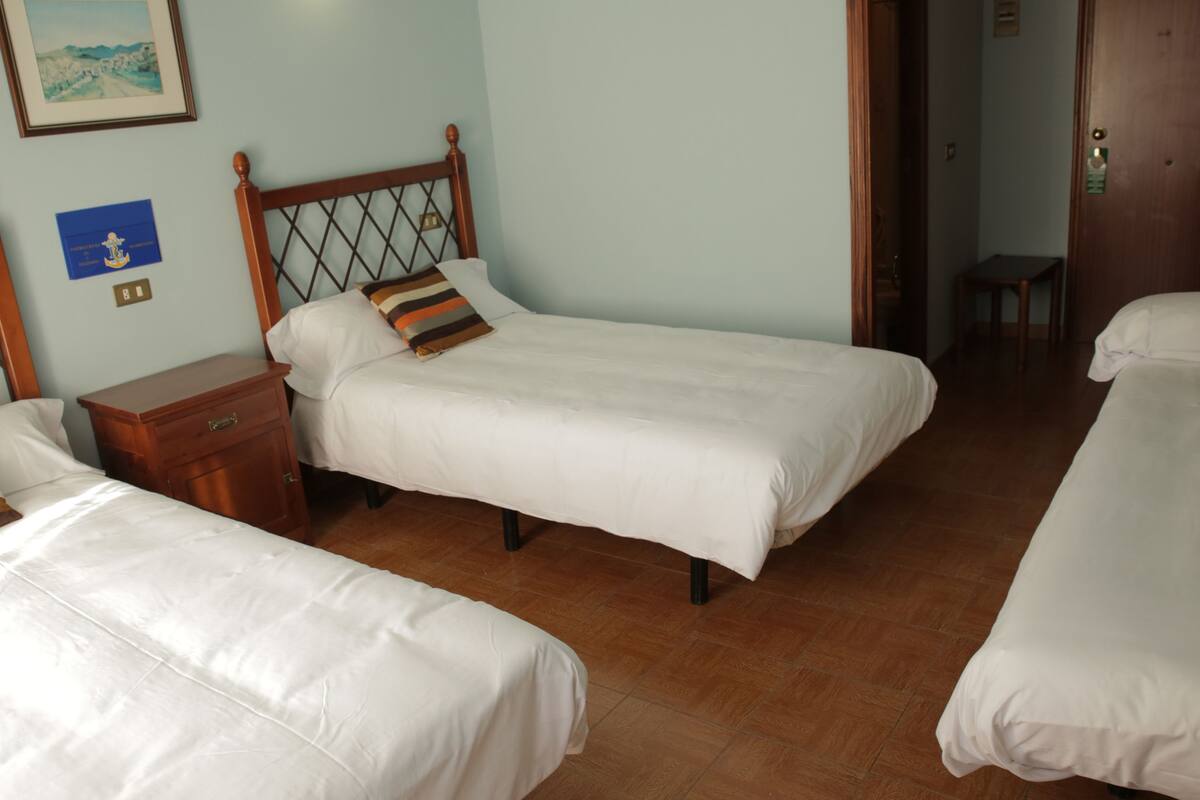 Quarto de hotel com três camas de solteiro, cabeceiras de madeira, uma mesinha de cabeceira e uma pequena mesa.