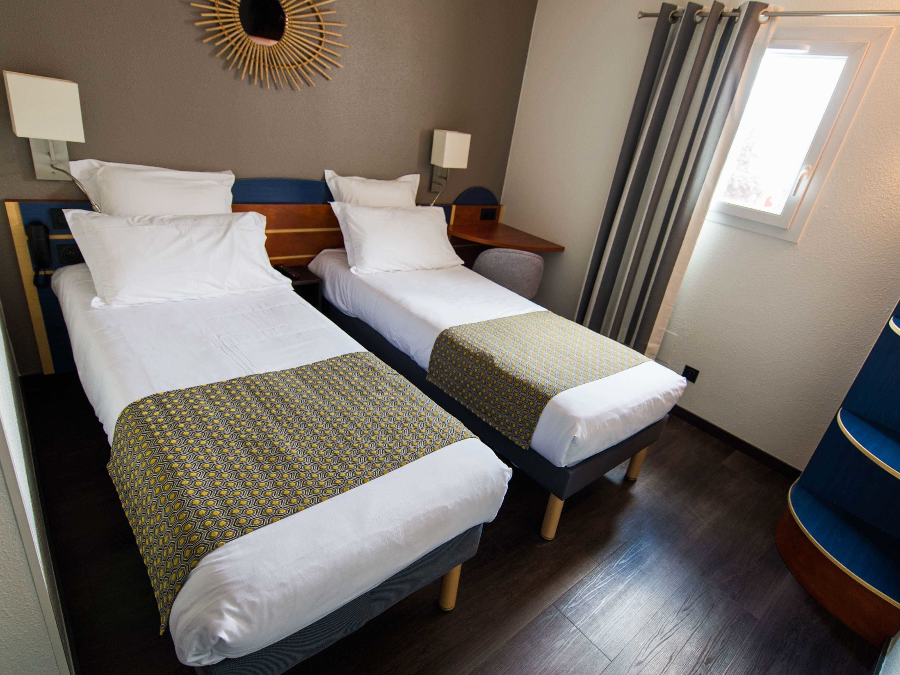 Quarto de hotel com duas camas de solteiro, uma pequena mesa de cabeceira com abajur, espelho fixado na parede e janela com cortinas.