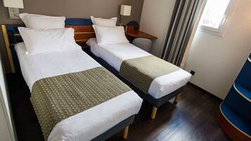 Quarto de hotel com duas camas de solteiro, uma pequena mesa de cabeceira com abajur, espelho fixado na parede e janela com cortinas.
