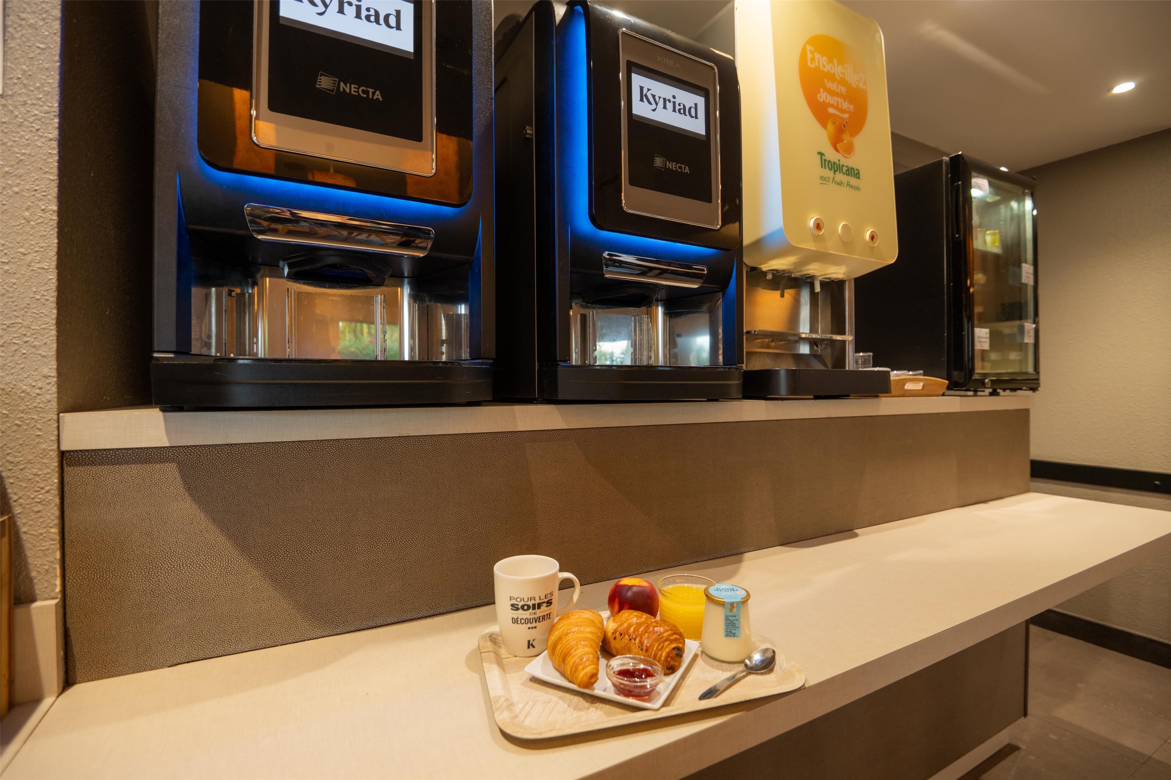 Bancada de pequeno-almoço com máquina de café, dispensador de sumo e bandeja com croissants, fruta e uma bebida.
