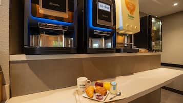 Bancada de pequeno-almoço com máquina de café, dispensador de sumo e bandeja com croissants, fruta e uma bebida.