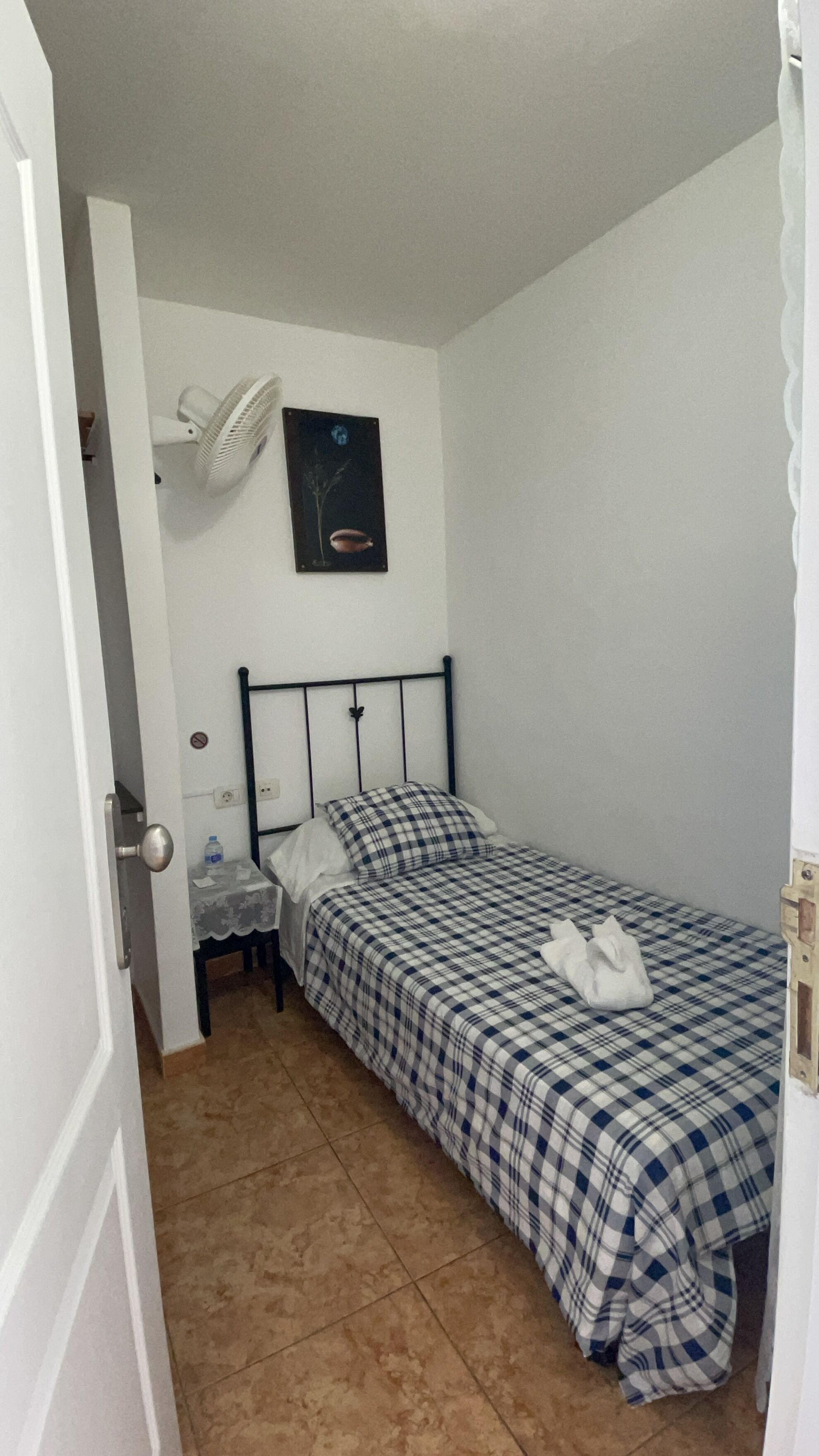 Um quarto pequeno com uma cama de solteiro, uma mesinha de cabeceira, um ventilador fixado na parede e um quadro emoldurado.