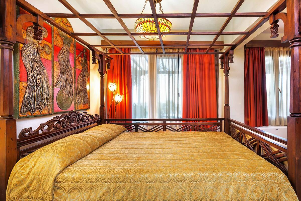 Uma cama grande com colcha estampada, cabeceira de madeira e vista para uma janela com cortinas vermelhas.