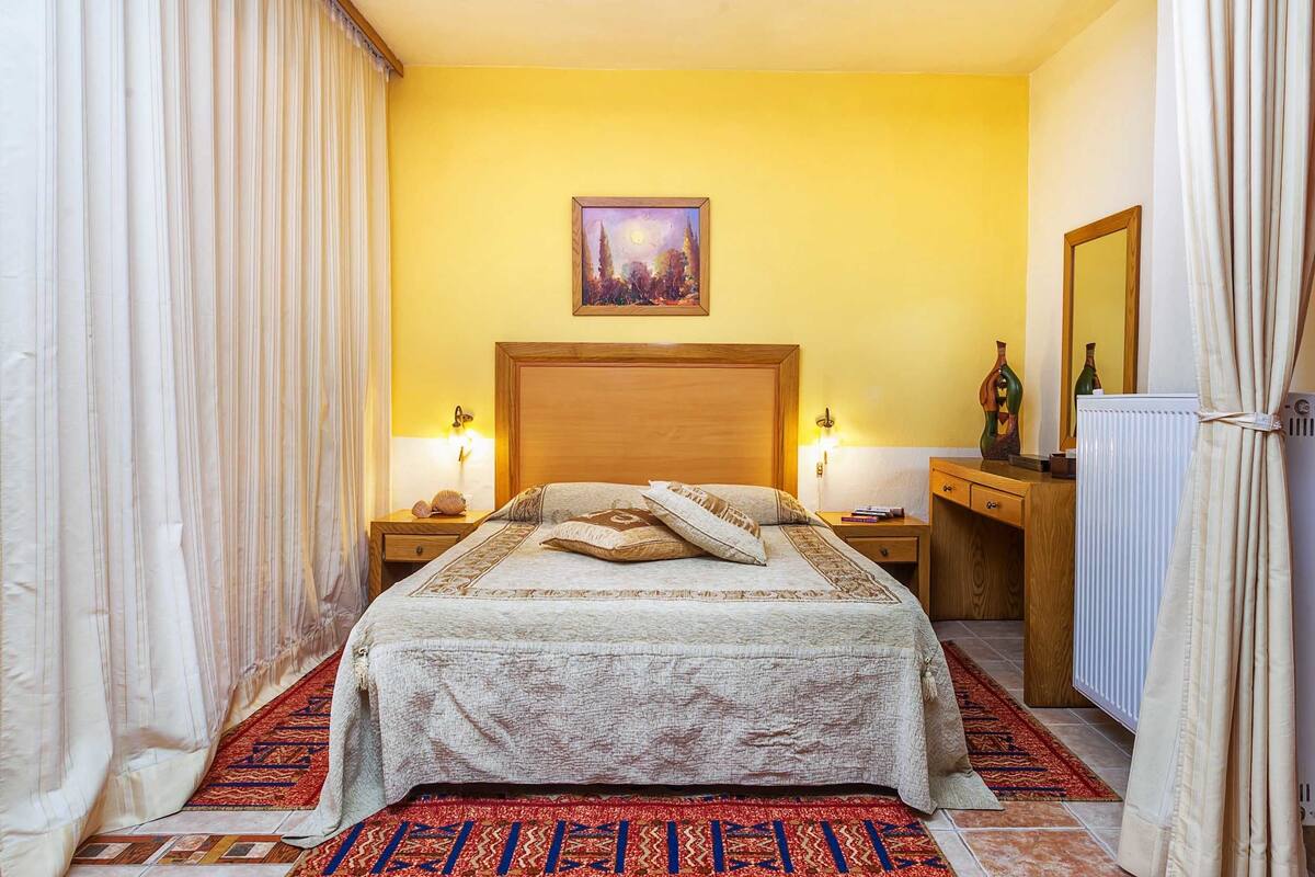 Quarto com uma cama grande, duas mesas de cabeceira com luminárias, cômoda de madeira, radiador e um quadro na parede.