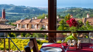Uma mesa de jantar com toalha vermelha, um vaso com flores vermelhas e uma vista panorâmica de casas e colinas ao fundo.
