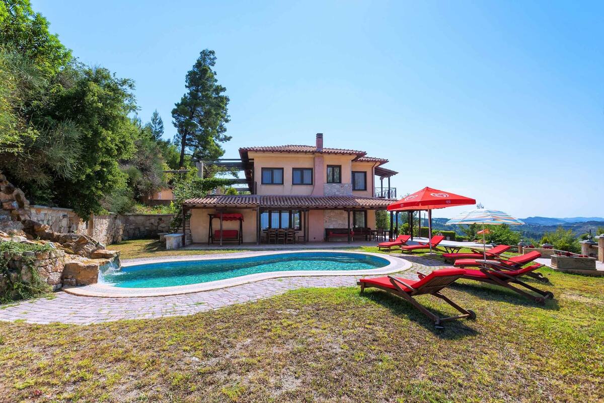 Uma casa de dois andares com piscina, cadeiras de descanso vermelhas e vista para as montanhas.