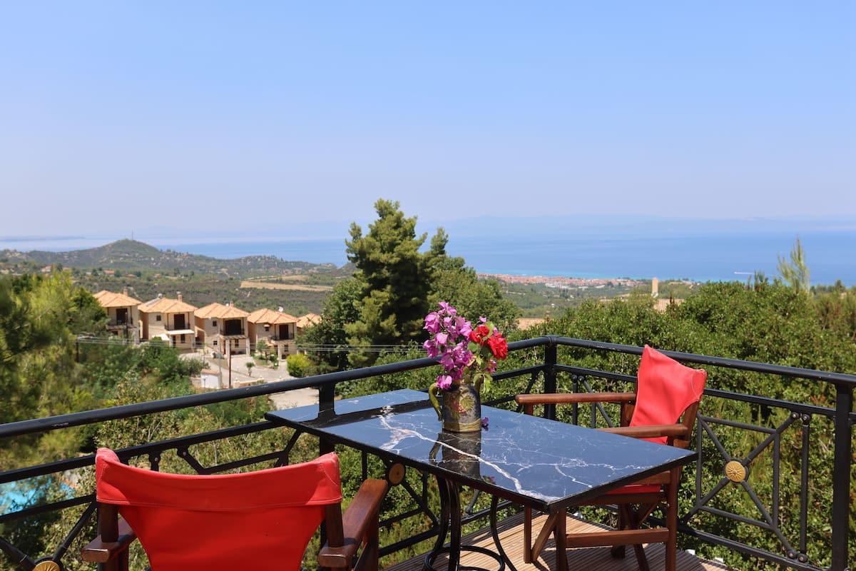 Um terraço com mesa e cadeiras, com vista para a paisagem costeira com edifícios e árvores.